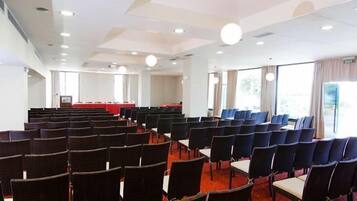 Sala de conferência com fileiras de cadeiras, palco com mesa vermelha e grandes janelas com cortinas.