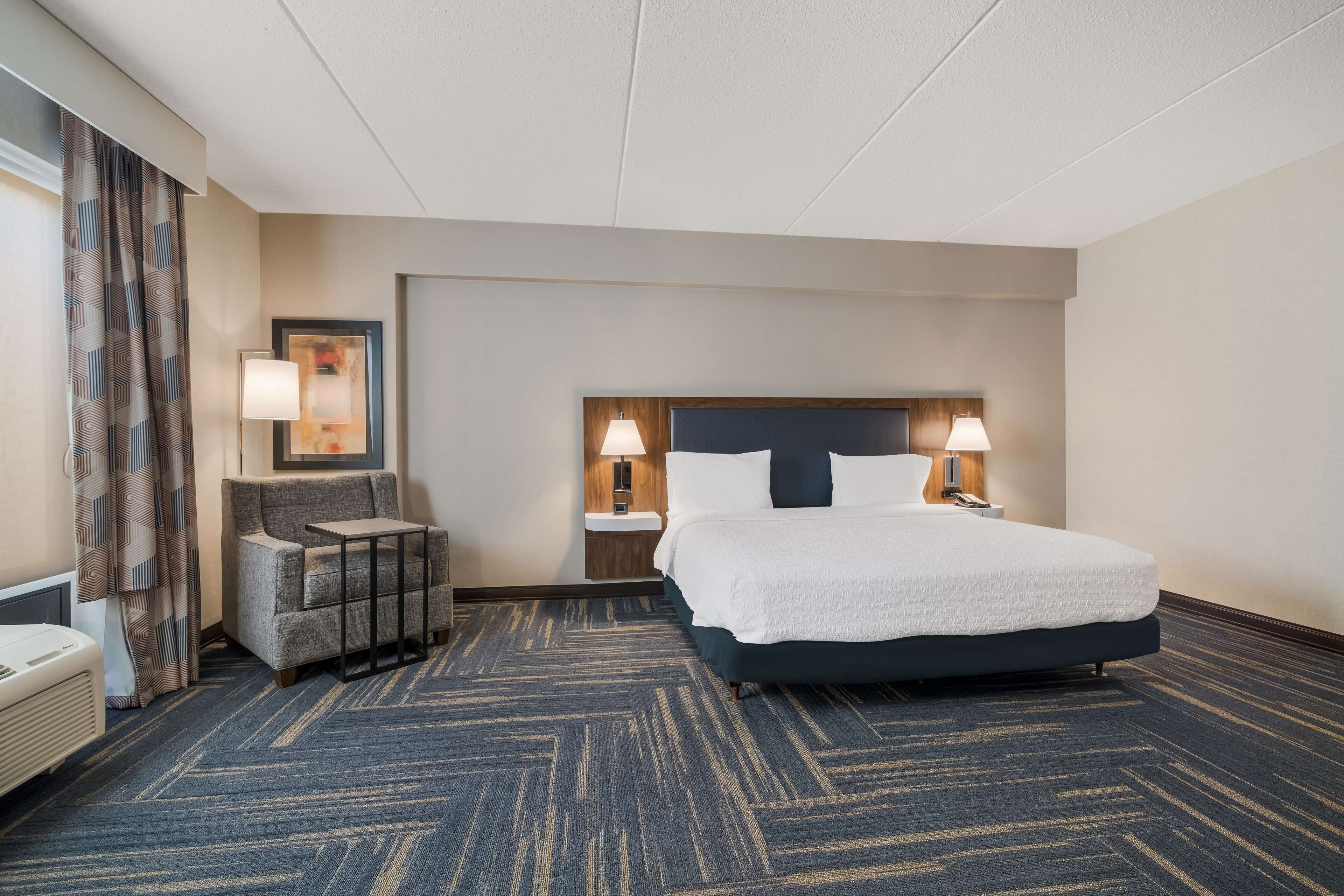 Quarto de hotel com uma cama grande, duas mesas de cabeceira com luminárias, uma cadeira, uma escrivaninha e uma janela ampla com cortinas.