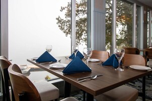 Mesa de jantar posta com guardanapos azuis, copos de vinho e talheres, com vista para árvores através de janelas amplas.