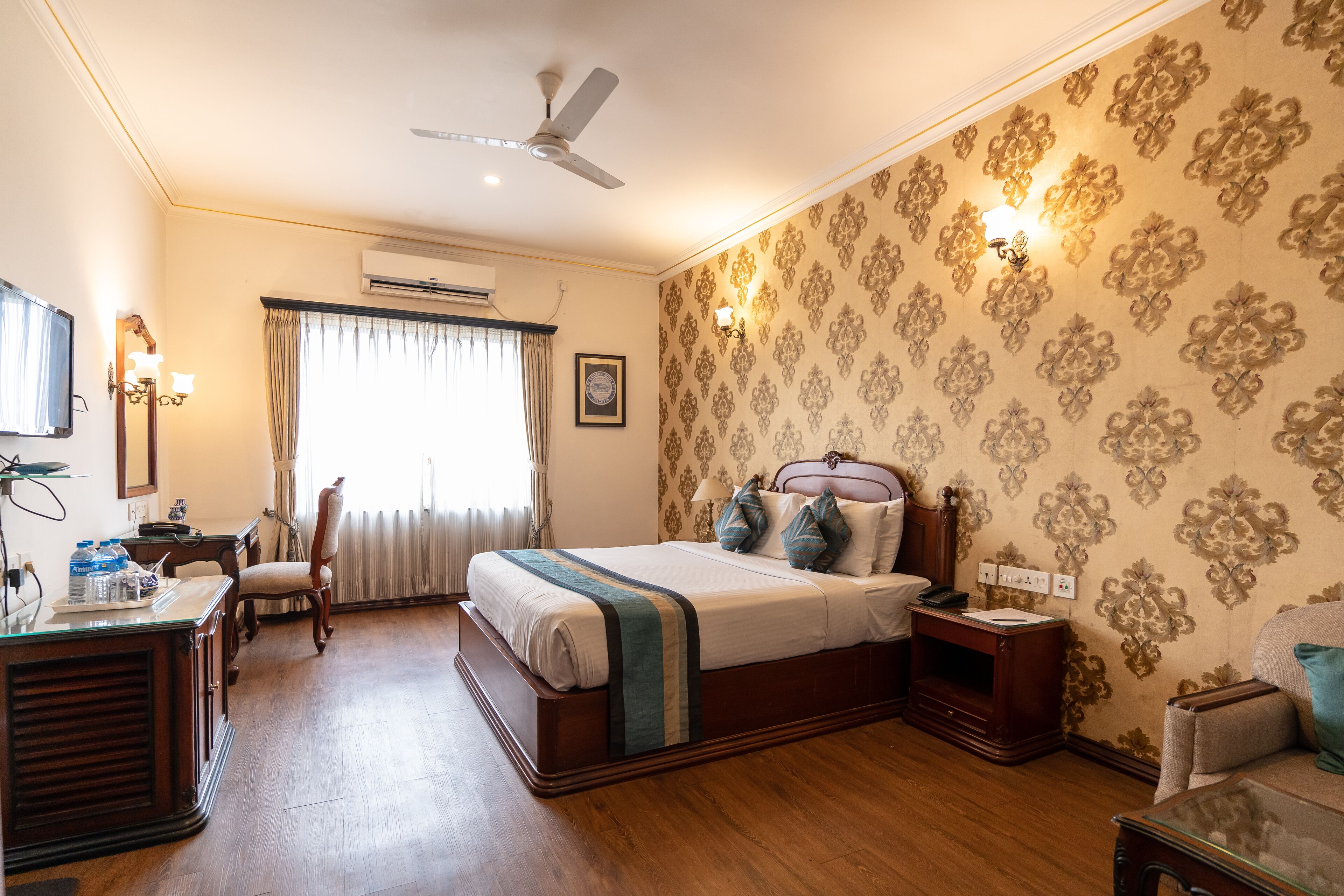 Quarto de hotel com cama, mesa de cabeceira, cadeira, escrivaninha e ventilador de teto.