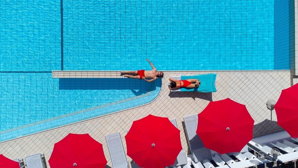 Piscina com paredes azulejadas, prancha de mergulho e guarda-sóis vermelhos.