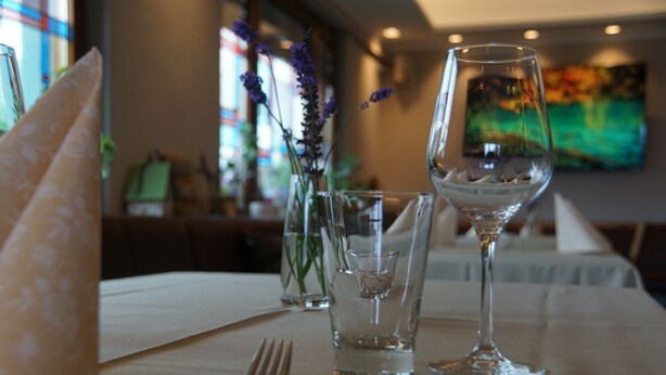 Mesa de jantar posta com uma taça de vinho, um copo pequeno e um garfo, com um vaso de lavanda ao fundo.