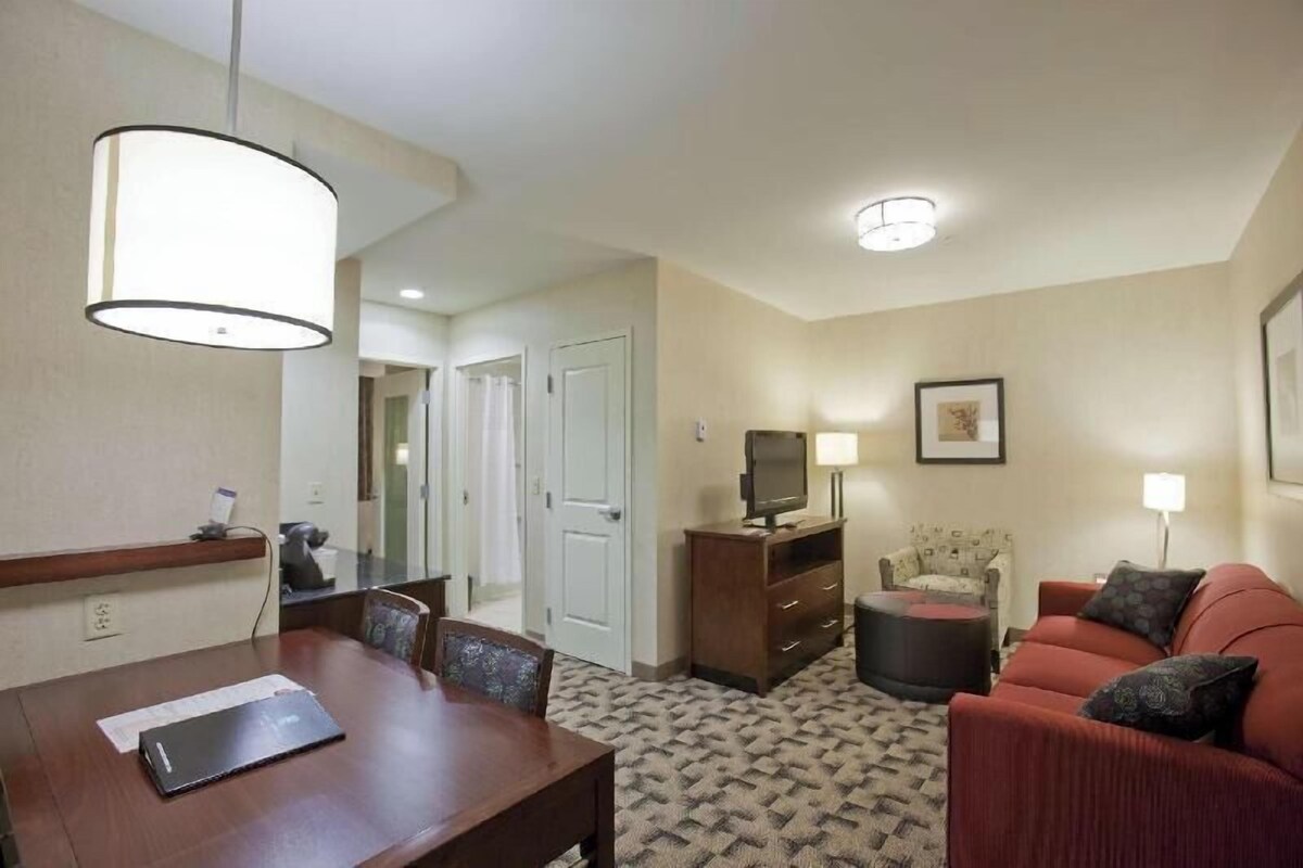 Quarto de hotel com uma mesa de jantar, um sofá, um aparador para TV e um abajur.