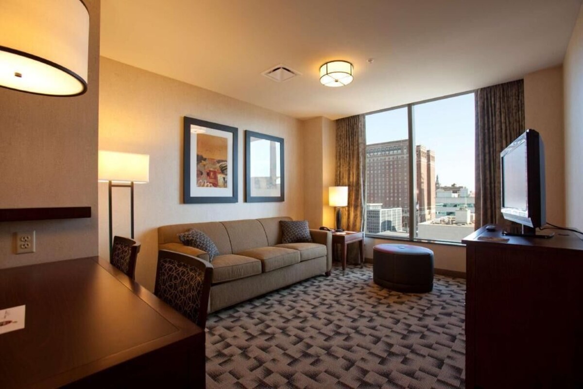 Quarto de hotel com sofá, televisão, escrivaninha, luminária e uma janela grande com vista para a cidade.