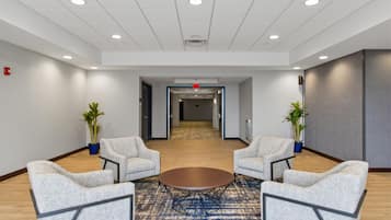 Um hall de hotel moderno com piso carpete, dois poltronas, uma mesa redonda e plantas em vasos.