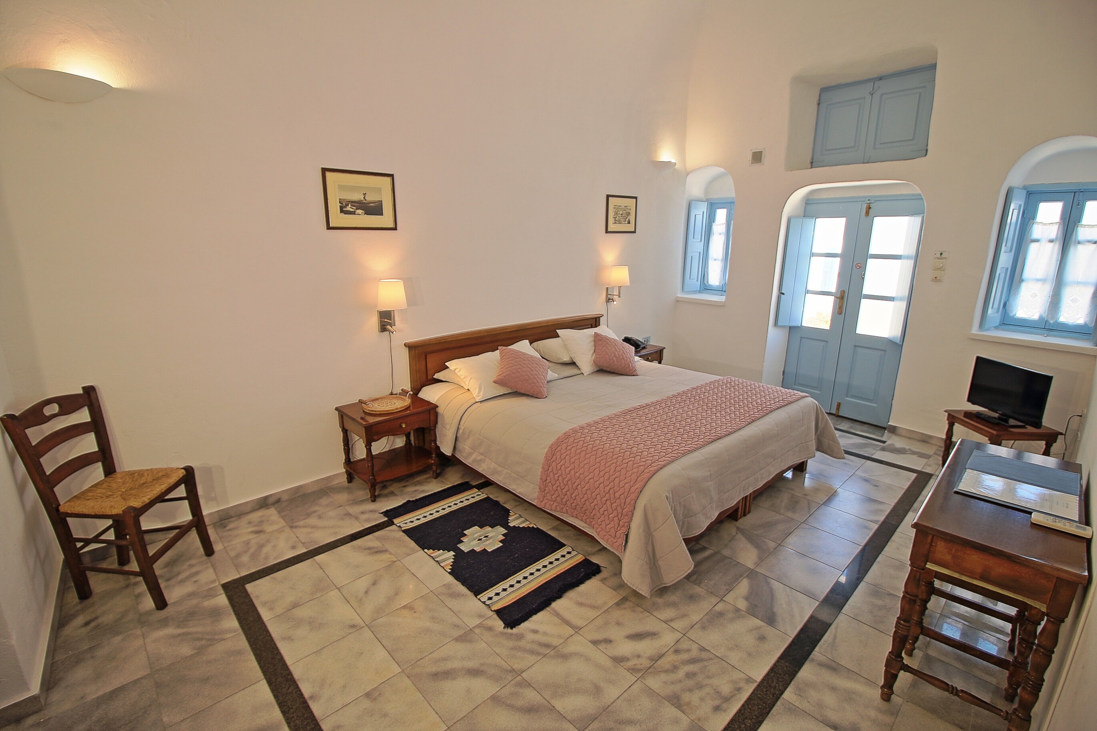 Quarto com cama, mesas de cabeceira, uma cadeira, escrivaninha, televisão e janela.
