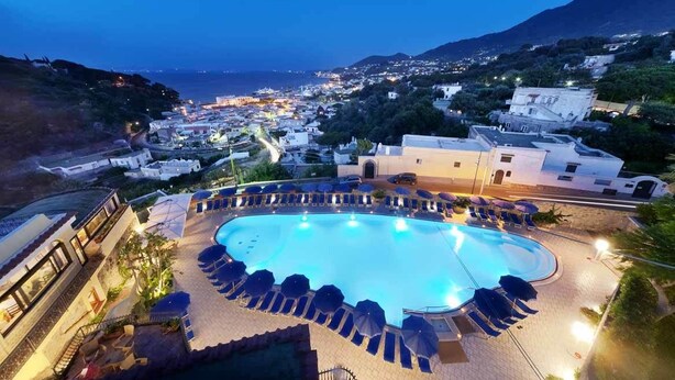 Área de piscina com espreguiçadeiras e guarda-sóis, cercada por edifícios com vista para o mar e uma montanha ao fundo.