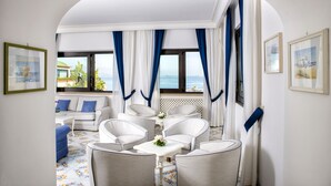 Quarto com sofá branco, poltronas brancas, uma mesinha e vista para o mar através de amplas janelas com cortinas azuis.
