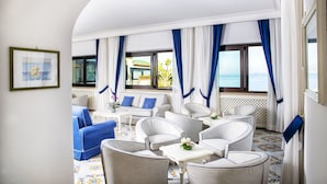 Quarto de hotel com esquema de cores branco e azul, com sofá, poltronas, uma mesa pequena e vista para o mar através de grandes janelas.
