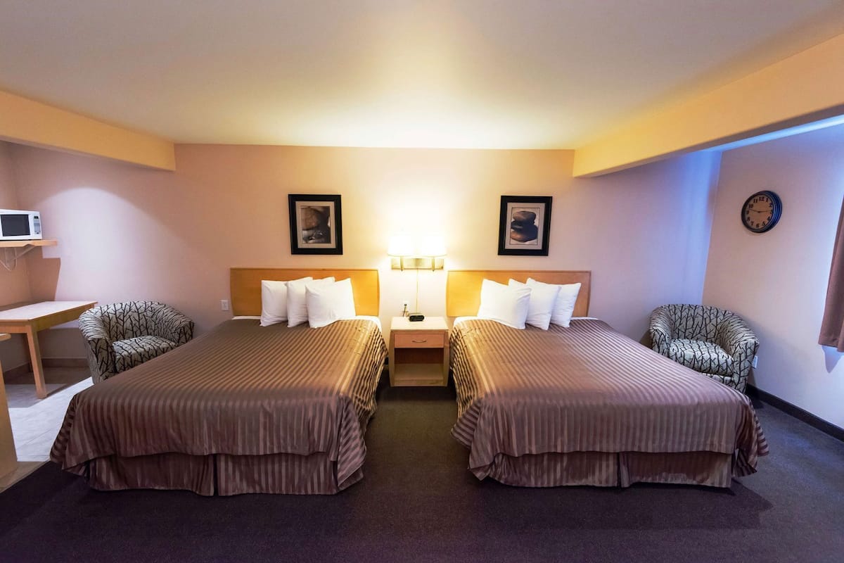 Quarto de hotel com duas camas, uma escrivaninha, uma cadeira, um abajur e um relógio na parede.