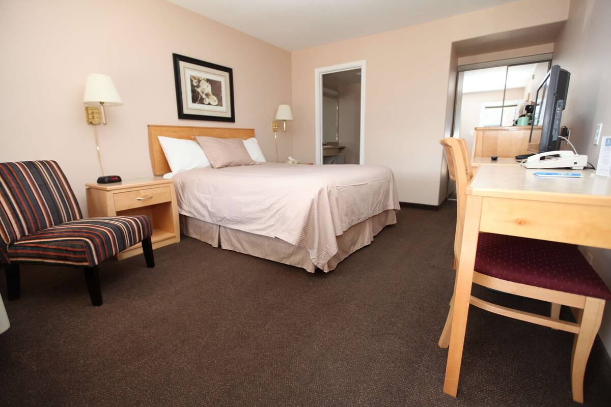 Quarto de hotel com cama, escrivaninha com computador, cadeira, mesa de cabeceira e luminária.