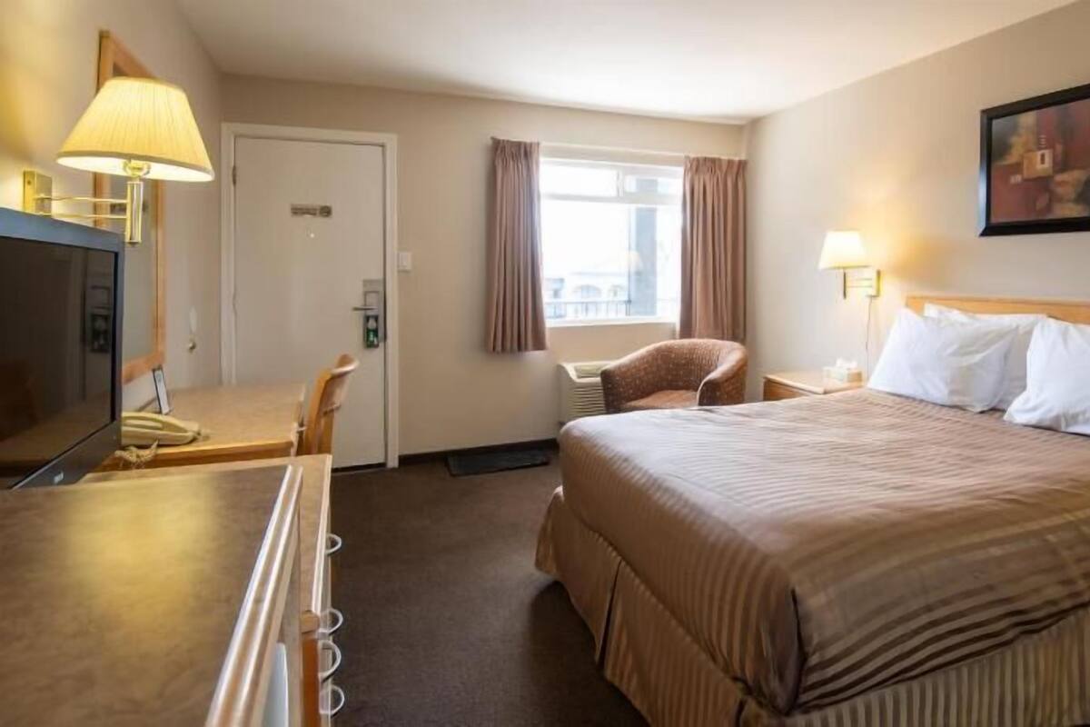 Quarto de hotel com cama, escrivaninha com cadeira, televisão e janela com cortinas.