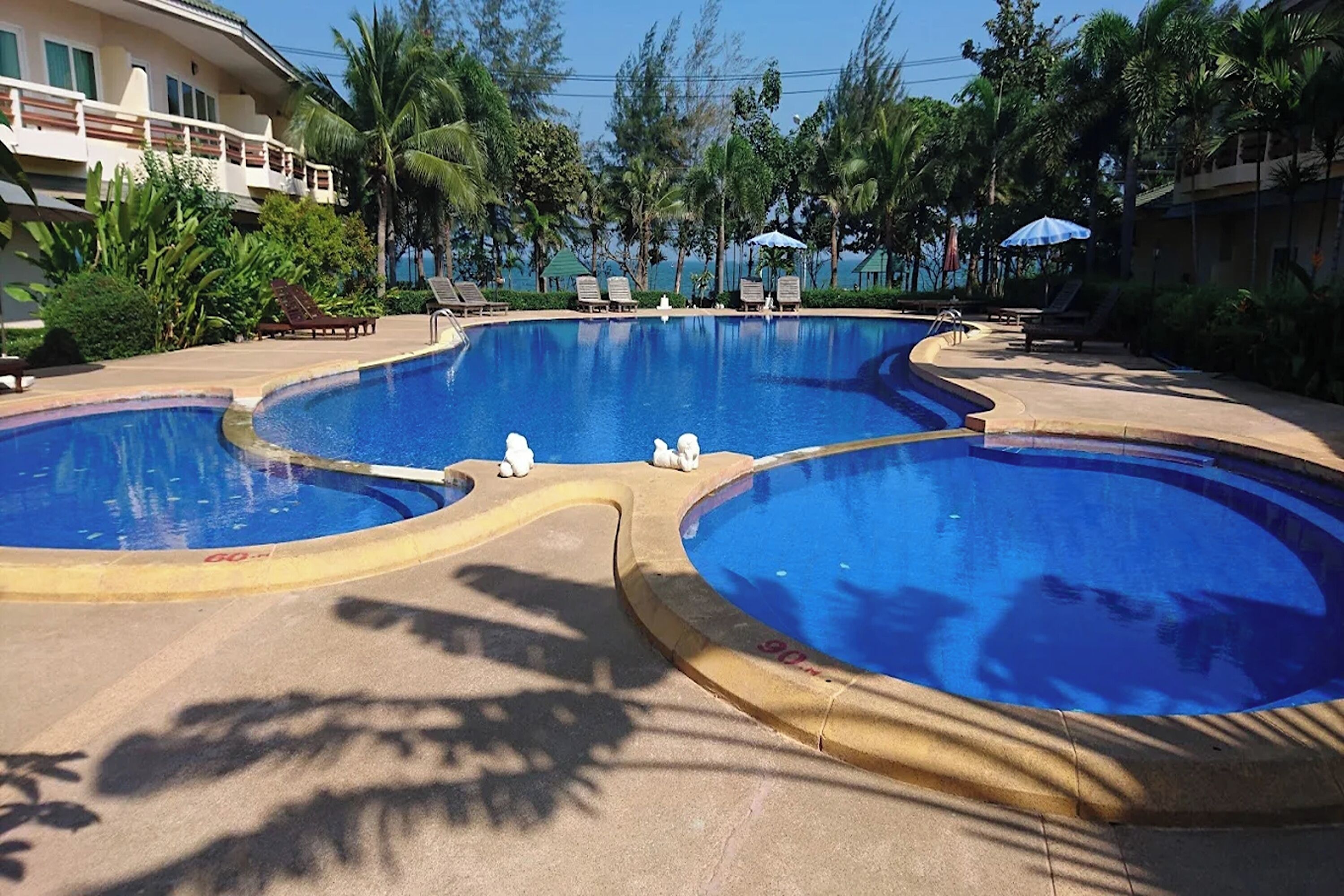 Uma grande piscina ao ar livre com várias áreas, cercada por espreguiçadeiras e guarda-sóis, com um edifício e palmeiras ao fundo.