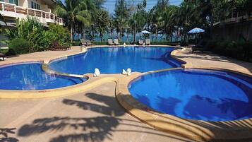 Uma grande piscina ao ar livre com várias áreas, cercada por espreguiçadeiras e guarda-sóis, com um edifício e palmeiras ao fundo.