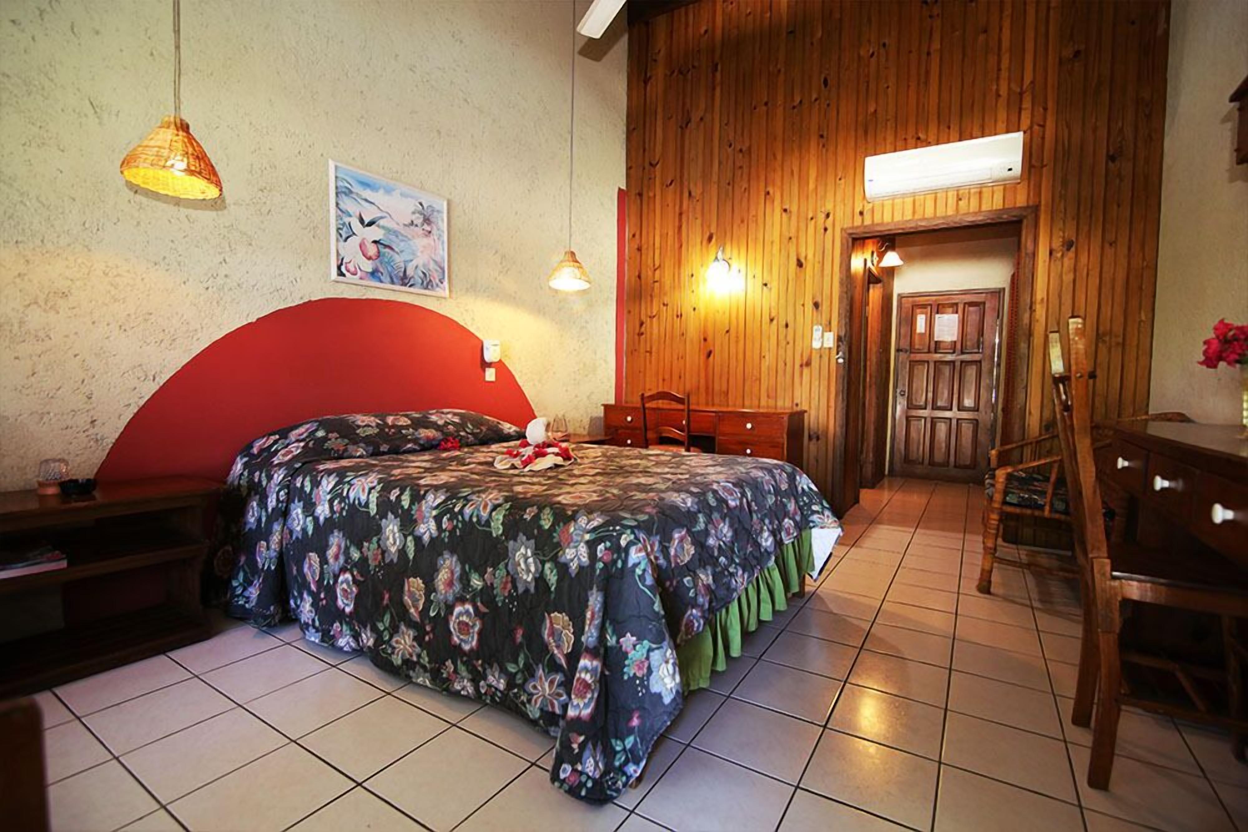 Quarto com colcha florida, paredes de madeira, cabeceira vermelha, escrivaninha de madeira e uma cadeira.