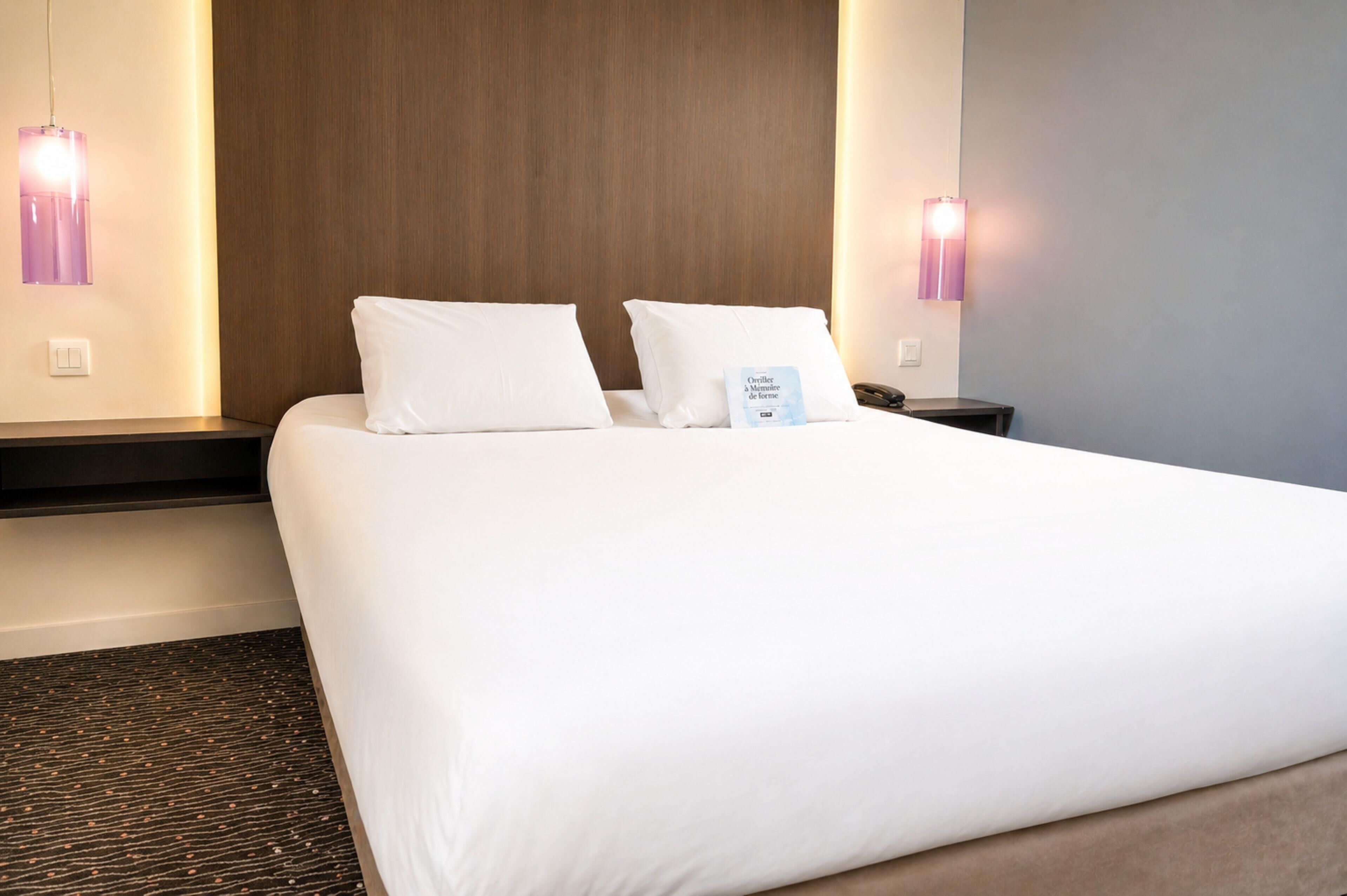 Quarto de hotel com uma cama grande, duas mesas de cabeceira, um painel de madeira na cabeceira e luminárias fixadas na parede.