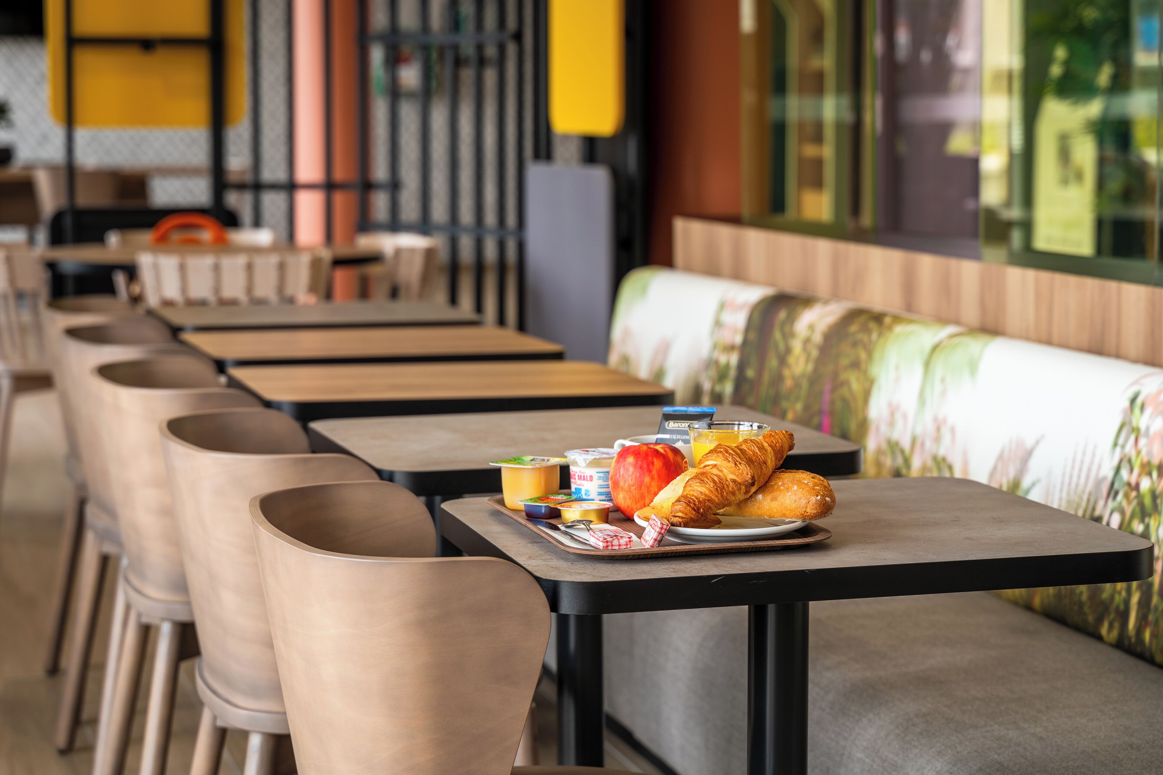 Um restaurante com várias mesas e cadeiras, uma bandeja com croissants, maçãs e bebidas sobre uma das mesas.