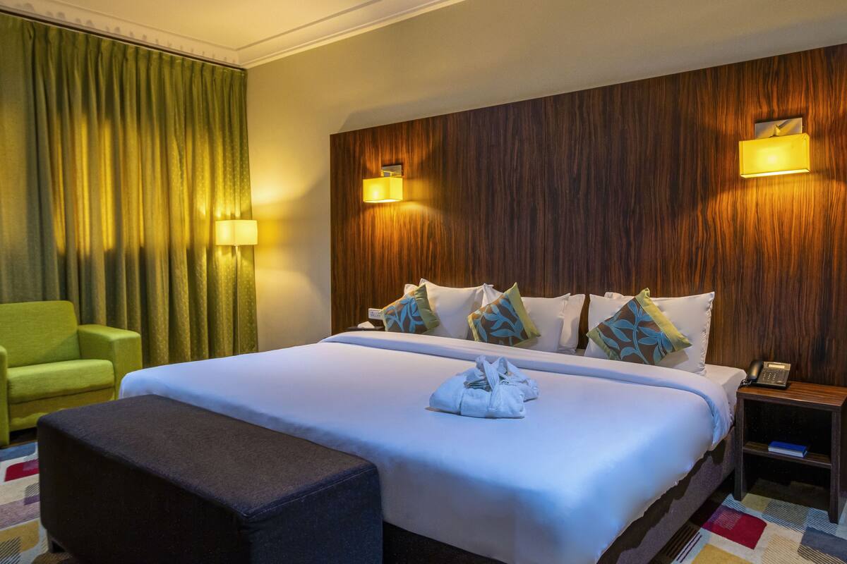 Quarto de hotel com uma cama grande, uma poltrona verde, uma mesinha e uma cabeceira de madeira.