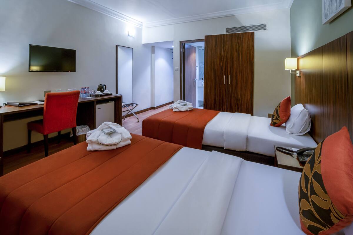 Quarto de hotel com duas camas, uma escrivaninha, uma cadeira, uma televisão e um guarda-roupa.