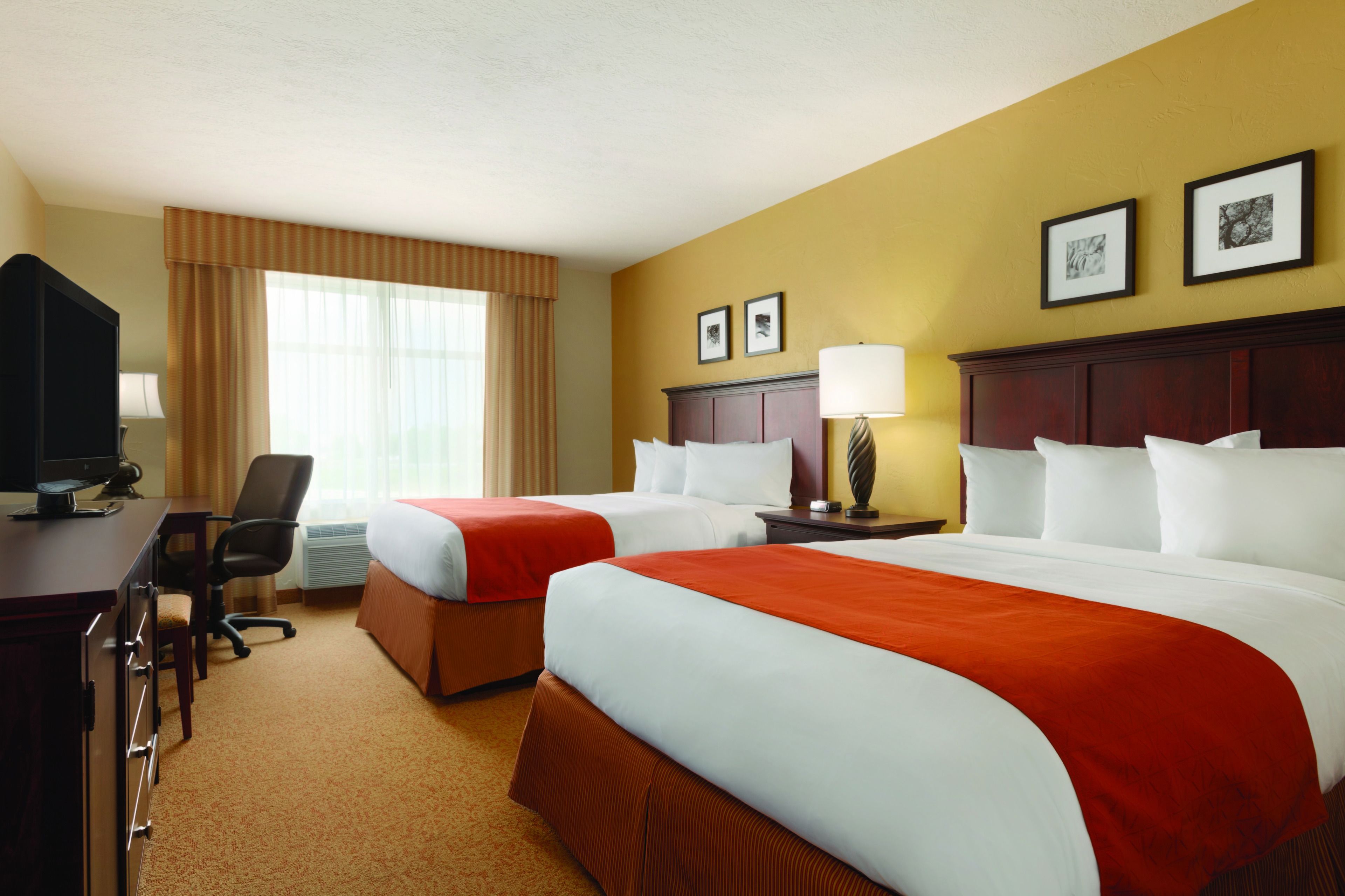 Quarto de hotel com duas camas, uma secretária com computador, uma cadeira e uma janela com cortinas.