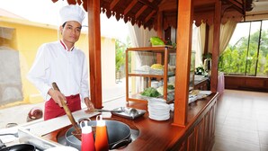 Um cozinheiro de uniforme branco e avental vermelho, em uma cozinha com bancada de madeira, fogão e diversos utensílios.