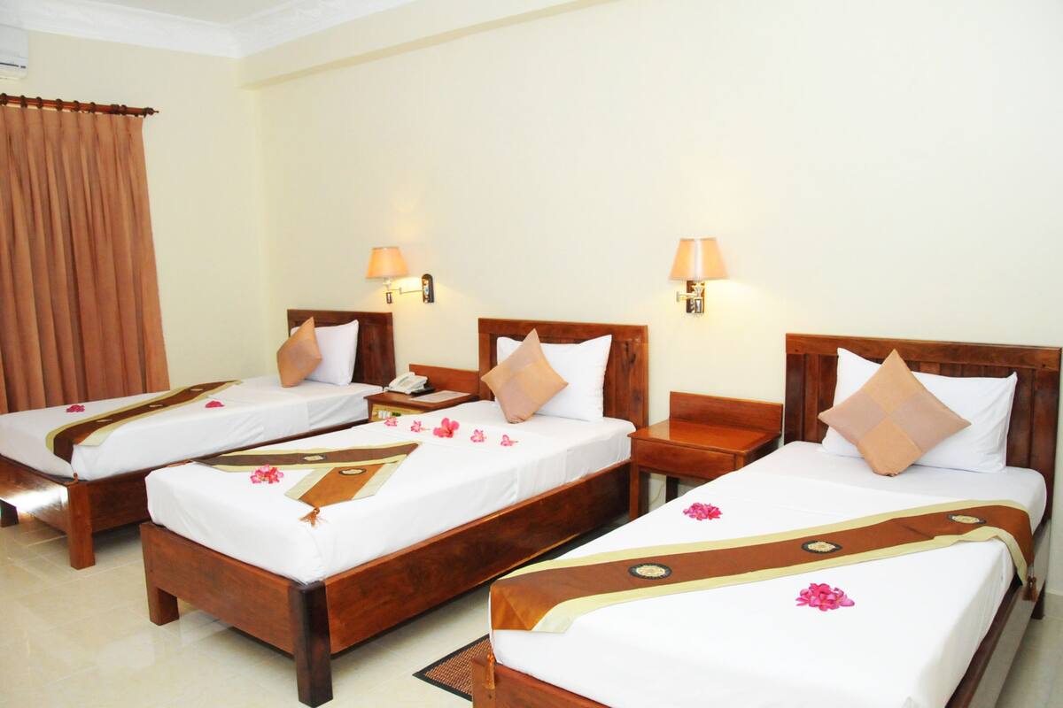 Quarto de hotel com três camas de solteiro, cada uma com cabeceira de madeira e roupa de cama branca. Há duas luminárias fixadas na parede, uma de cada lado das camas.
