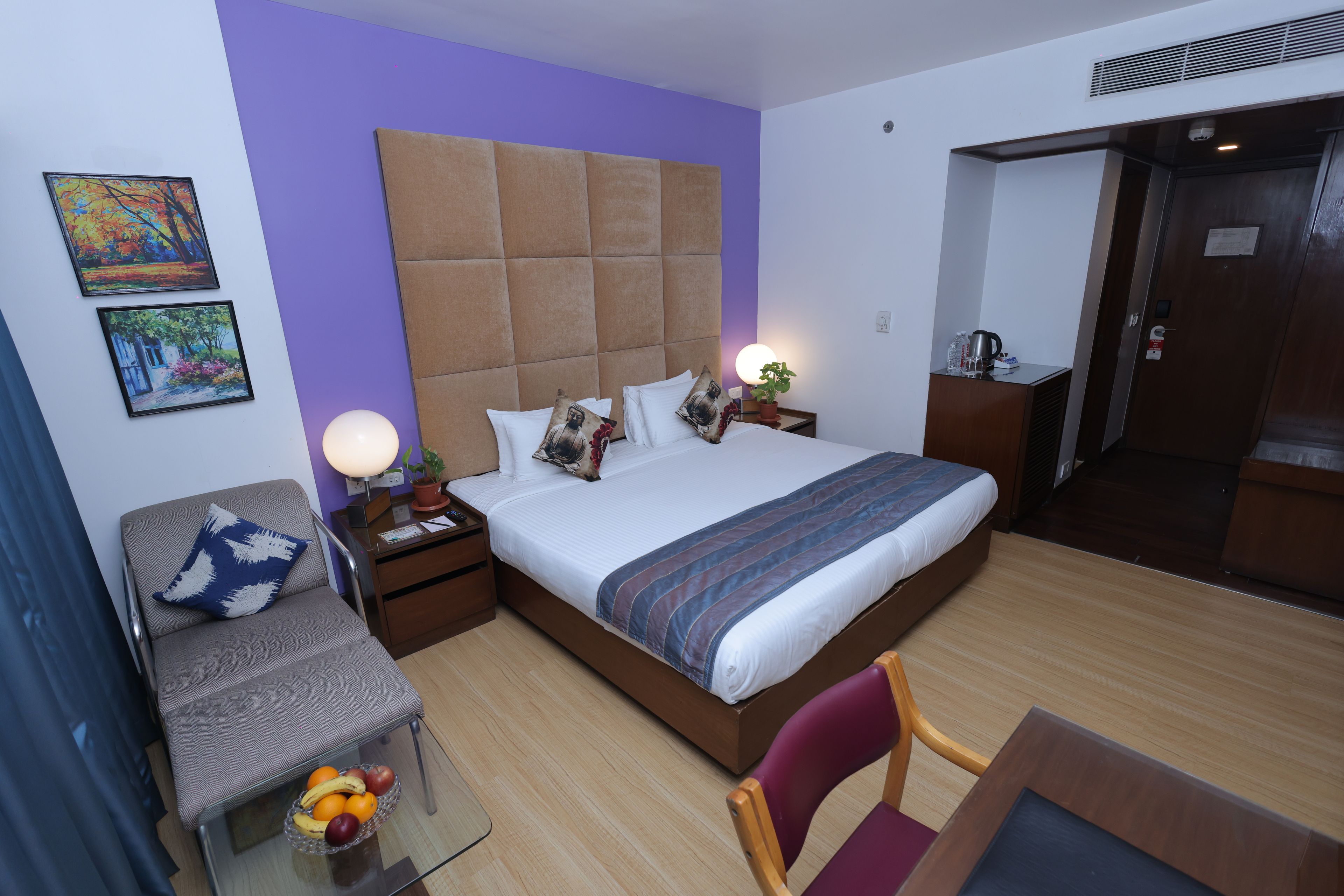 Quarto de hotel com cama, mesas de cabeceira, escrivaninha, cadeira, tigela de frutas e porta.