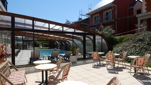 Um pátio externo com mesas redondas e cadeiras de vime, piscina com cobertura de vidro e um edifício vermelho ao fundo.