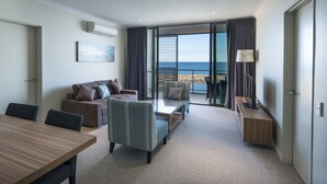 Uma sala de estar moderna com uma mesa de jantar de madeira, um sofá, uma poltrona listrada e uma varanda com vista para o mar.