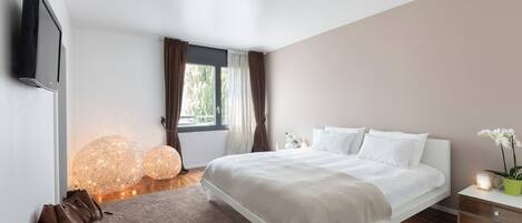 Quarto moderno com uma cama grande, televisão de tela plana na parede, janela com cortinas e mesa de cabeceira com abajur e planta.