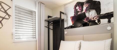 Quarto de hotel com cama, janela com persianas, um cabideiro e uma grande moldura na parede.