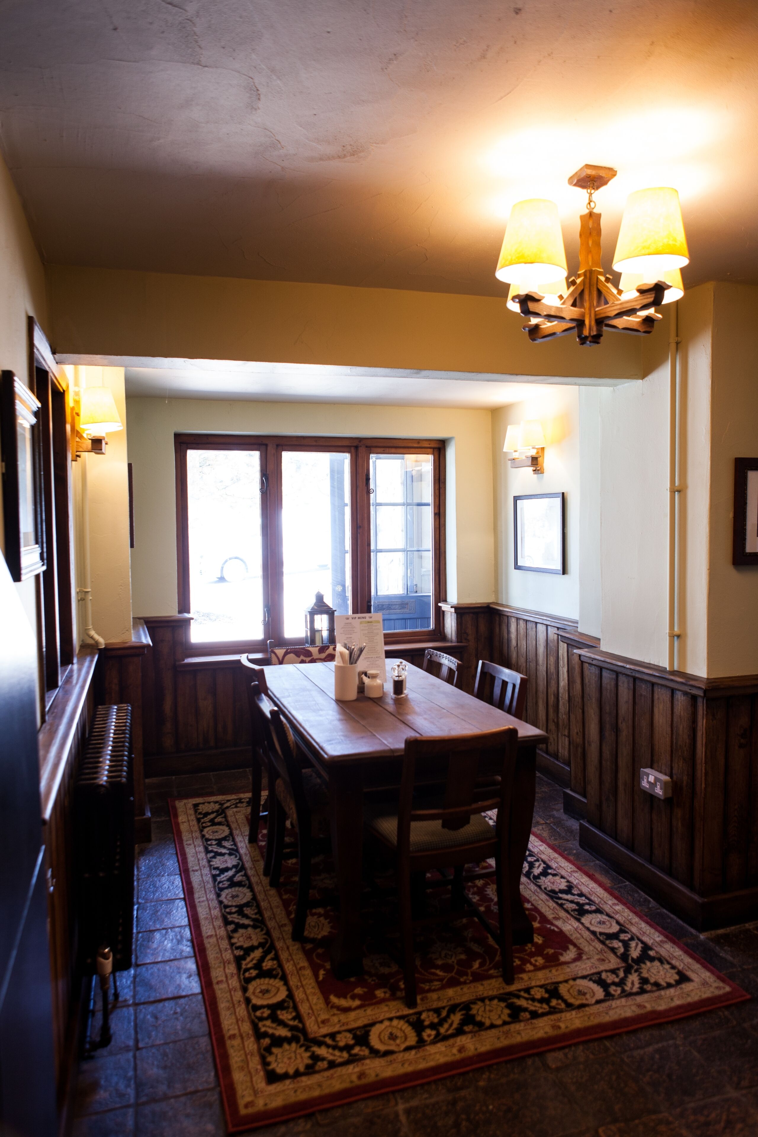 Uma sala de jantar com mesa de madeira, cadeiras e um tapete estampado. Há duas janelas com cortinas, um lustre e luminárias na parede.
