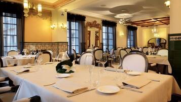Uma sala de jantar formal com várias mesas preparadas para o serviço, elegantes lustres e amplas janelas.