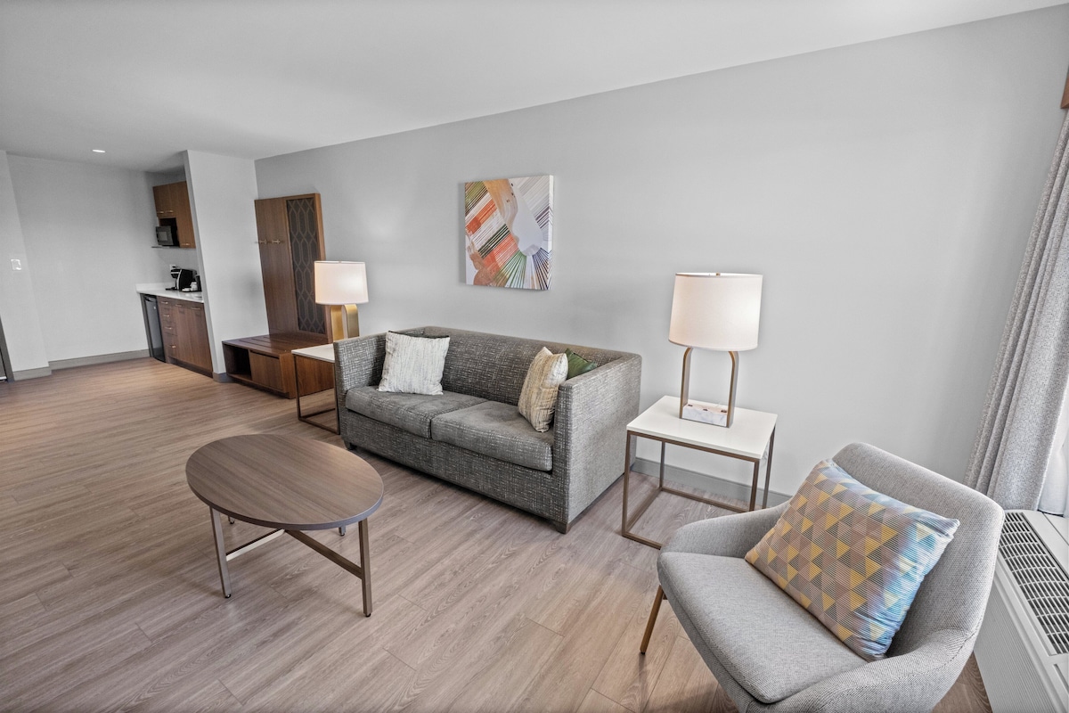 Uma sala de estar moderna com um sofá cinzento, uma mesa de centro redonda e uma poltrona cinzenta. Há um quadro na parede, uma pequena mesa com abajur e uma cozinha compacta ao fundo.