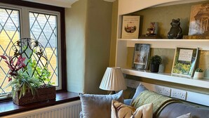 Uma sala de estar aconchegante com um sofá cinzento, um radiador, uma janela com uma planta e uma estante com livros e objetos decorativos.