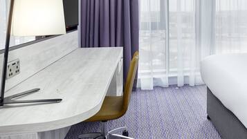Quarto de hotel com escrivaninha de madeira, uma cadeira, uma cama e vista da cidade através de cortinas finas.
