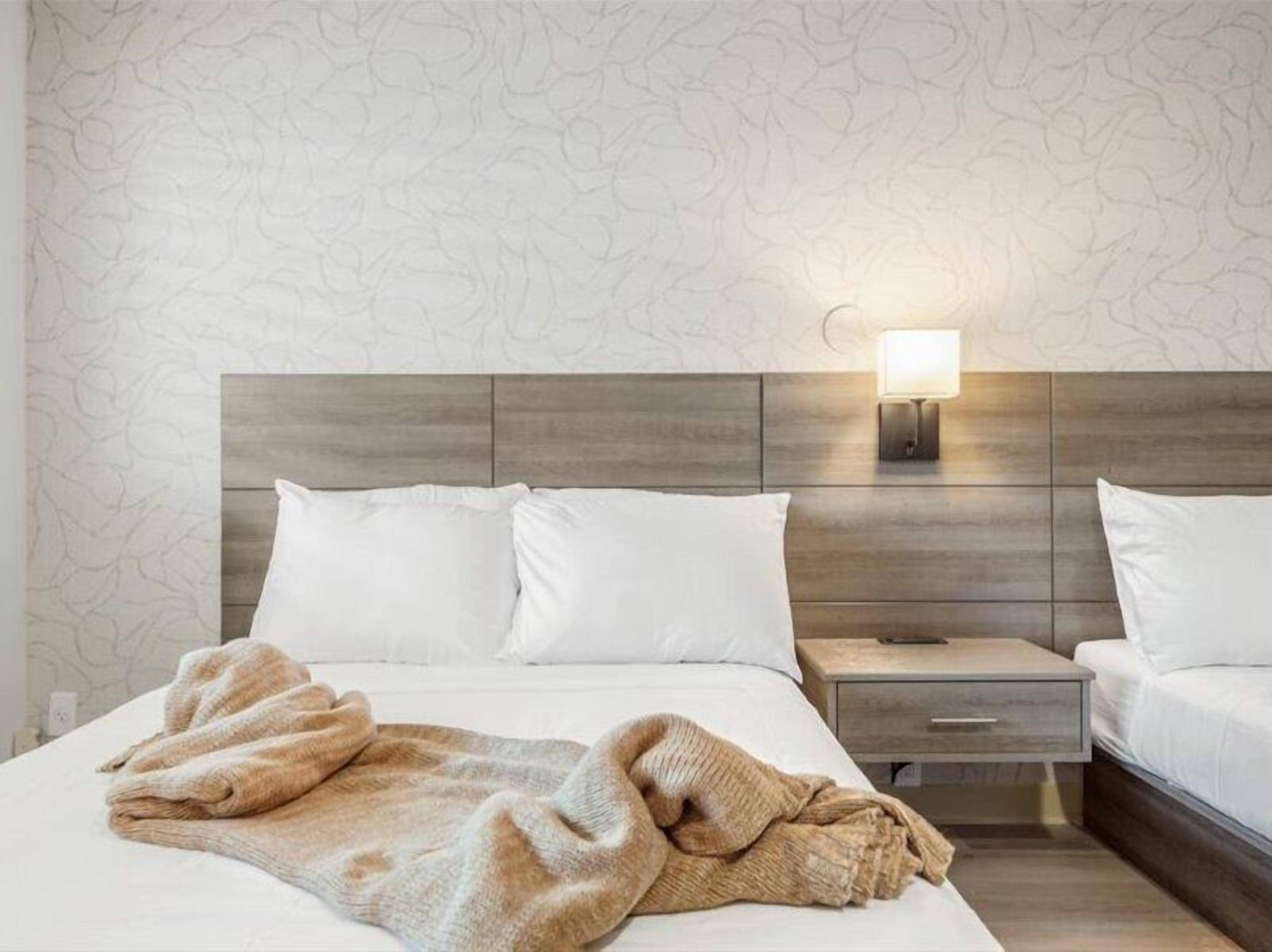 Quarto de hotel com cama, duas almofadas, manta, mesa de cabeceira e luminária na parede.