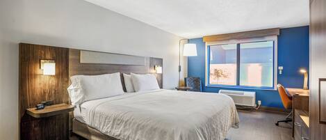 Quarto de hotel moderno com uma cama grande, mesa de cabeceira com abajur, escrivaninha com cadeira e janela com vista.