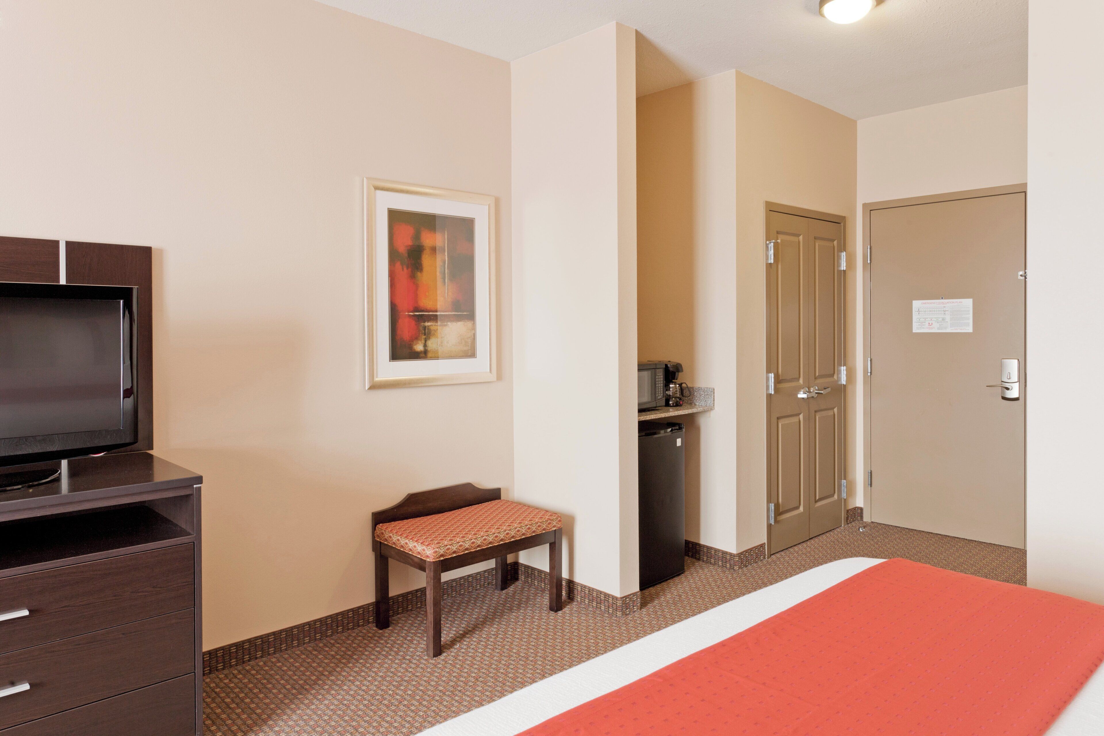 Quarto de hotel com cama, televisão, escrivaninha com cadeira, micro-ondas e porta com placa.