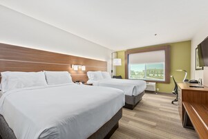 Quarto de hotel com duas camas, uma escrivaninha, uma cadeira e uma janela grande com vista para a cidade.