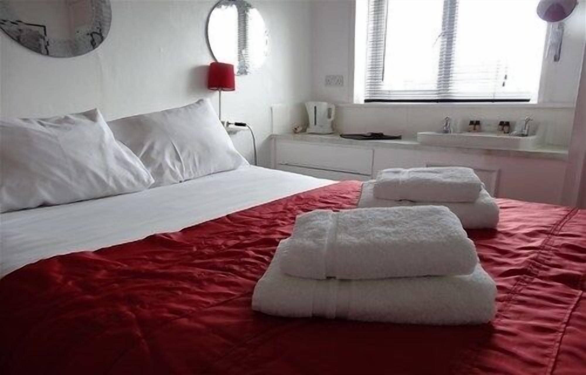 Cama bem arrumada com lençóis brancos e manta vermelha, mesa de cabeceira com abajur vermelho e janela com persianas.