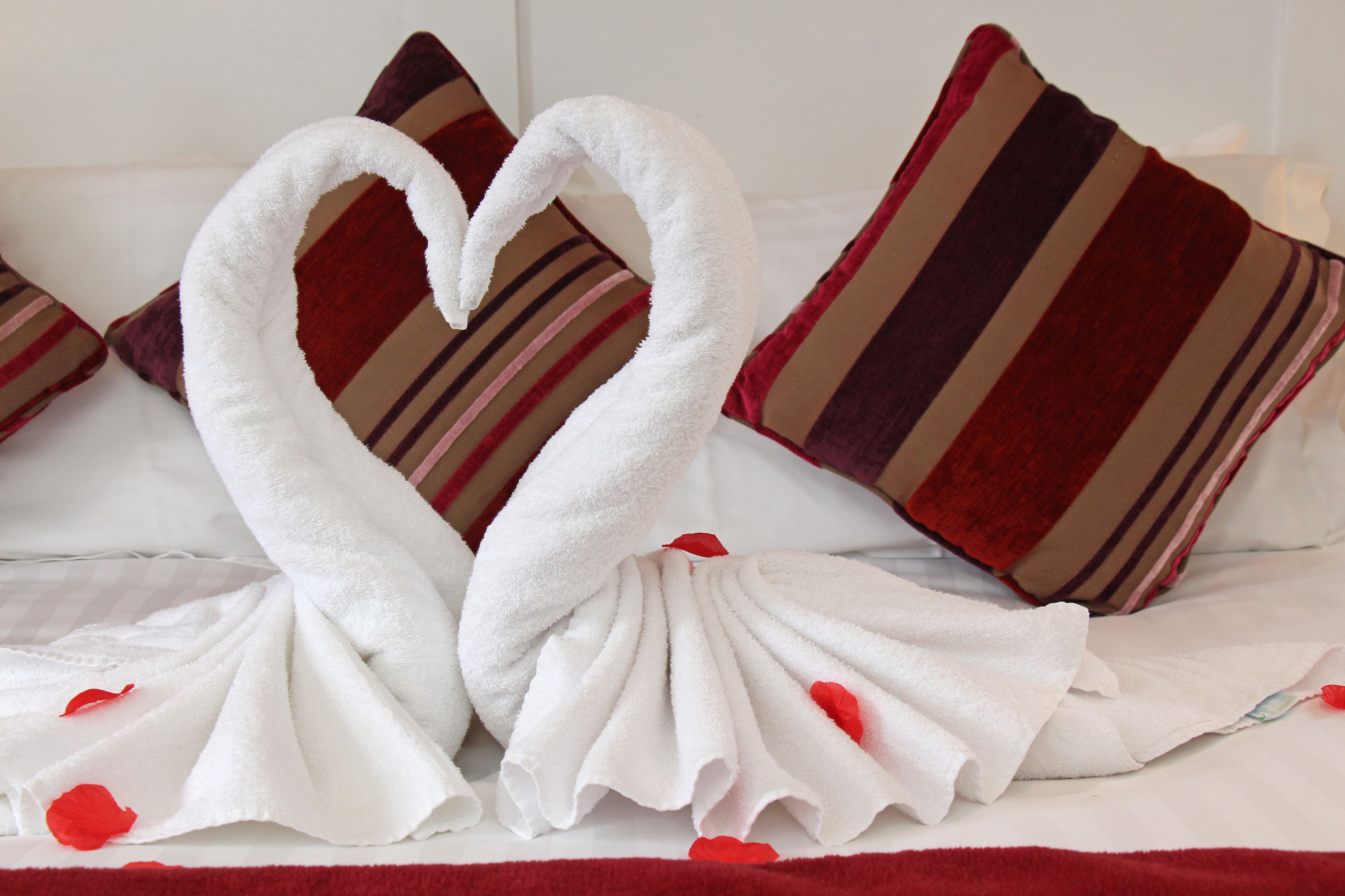 Cama com toalhas brancas dispostas em forma de dois cisnes, pétalas de rosas vermelhas espalhadas sobre a cama e almofadas listradas.
