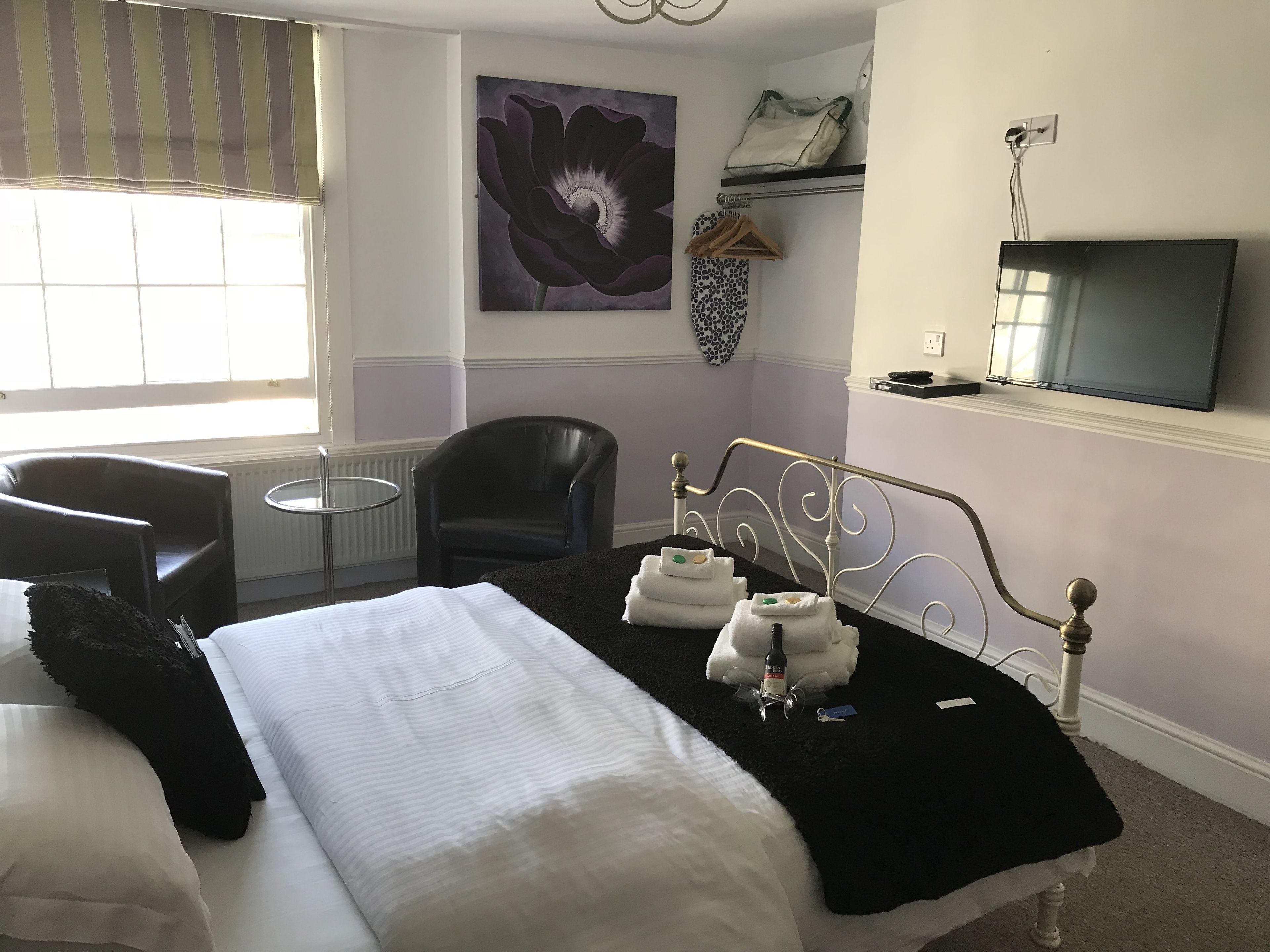 Quarto de hotel com uma cama, uma cadeira, uma mesinha, uma televisão e um quadro na parede.