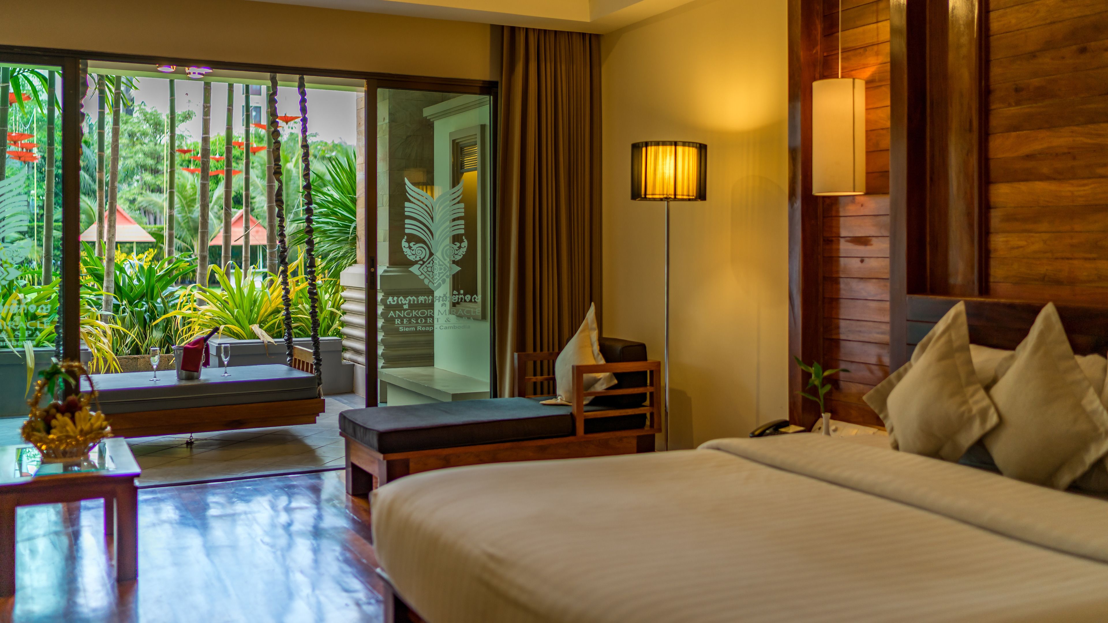 Quarto de hotel com cama, escrivaninha, cadeira e vista para um jardim exuberante através de uma porta de vidro.