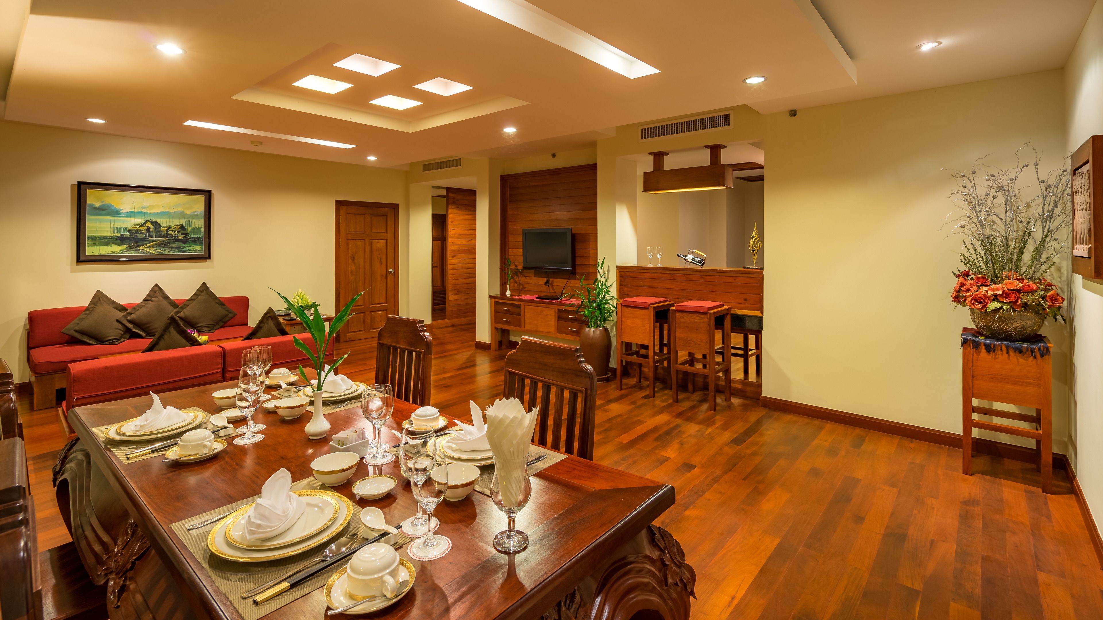 Uma área de refeições com uma mesa de madeira posta para uma refeição, um sofá vermelho e uma televisão ao fundo.