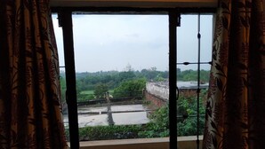 Vista pela janela com cortinas, mostrando um edifício grande à distância, rodeado por vegetação.