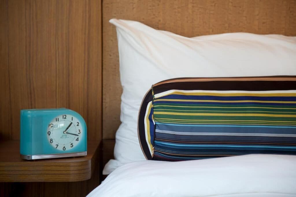 Uma cama com travesseiro branco e um travesseiro listrado, um relógio azul-turquesa em uma mesa de cabeceira de madeira.
