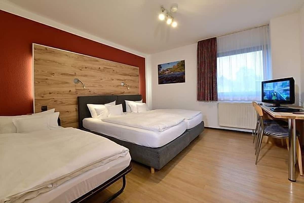 Quarto de hotel com duas camas, cabeceira de madeira, escrivaninha com cadeira, televisão e janela com cortinas.