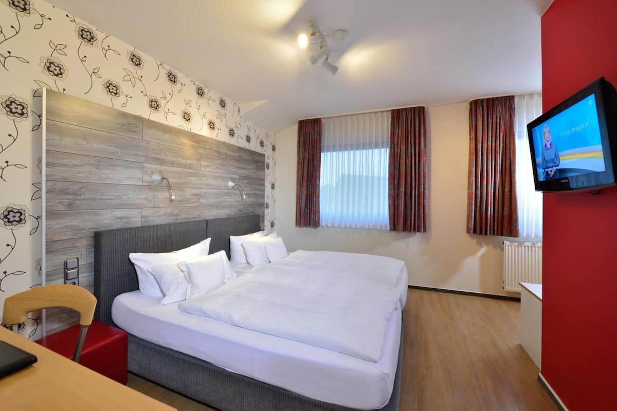 Quarto de hotel com uma cama grande, cabeceira de madeira, televisão montada na parede e uma janela com cortinas.