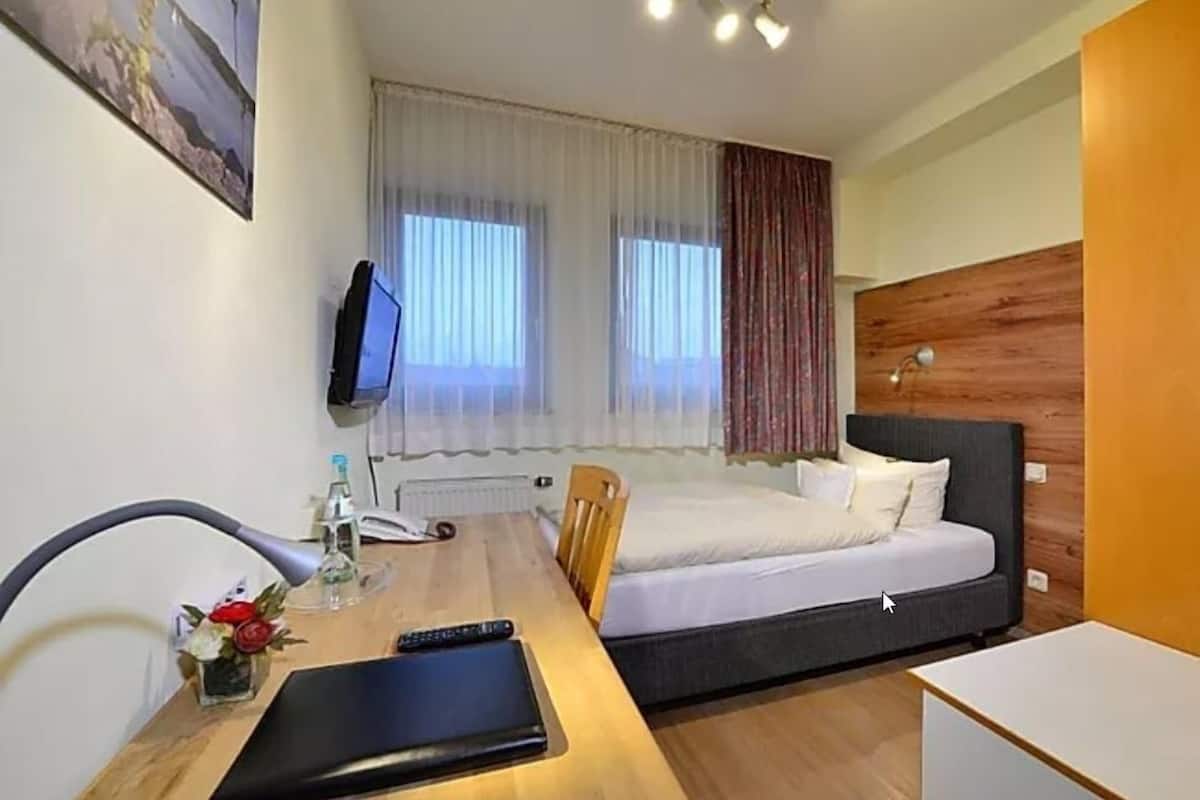 Quarto de hotel com uma cama, uma escrivaninha com cadeira, televisão e janela com cortinas.